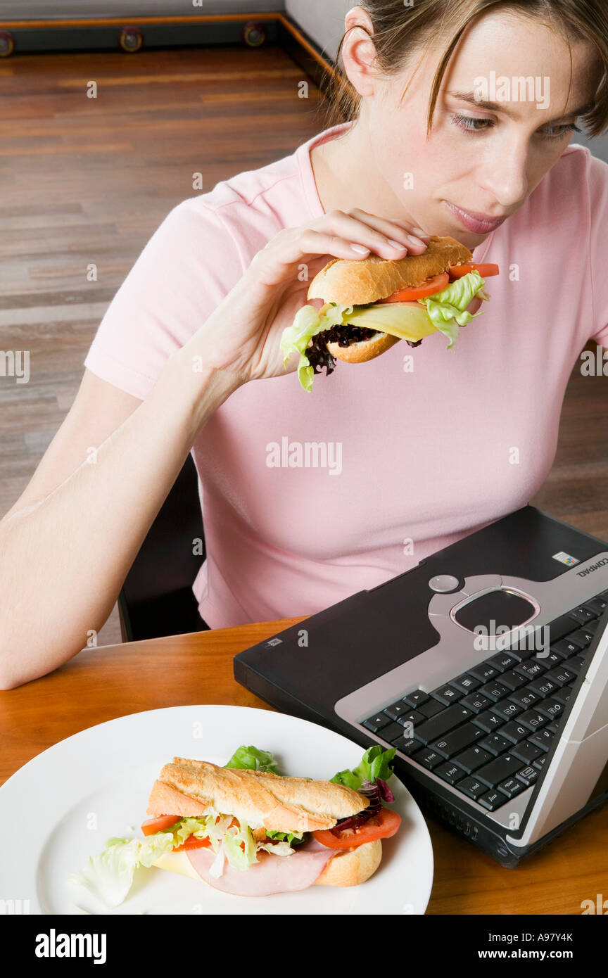 Young woman eating sandwich while working at computer FoodCollection ...