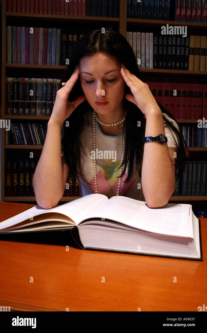 Young woman stressed while reading a large book at a desk in a library ...