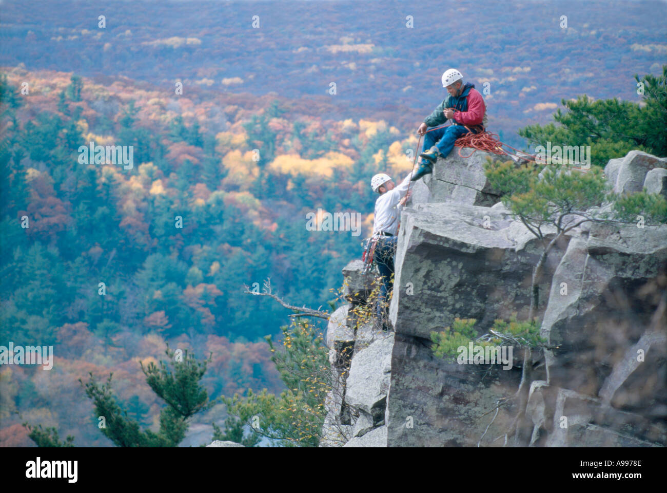 Rockclimber sits on a rock ledge assisting his companion up the cliff ...