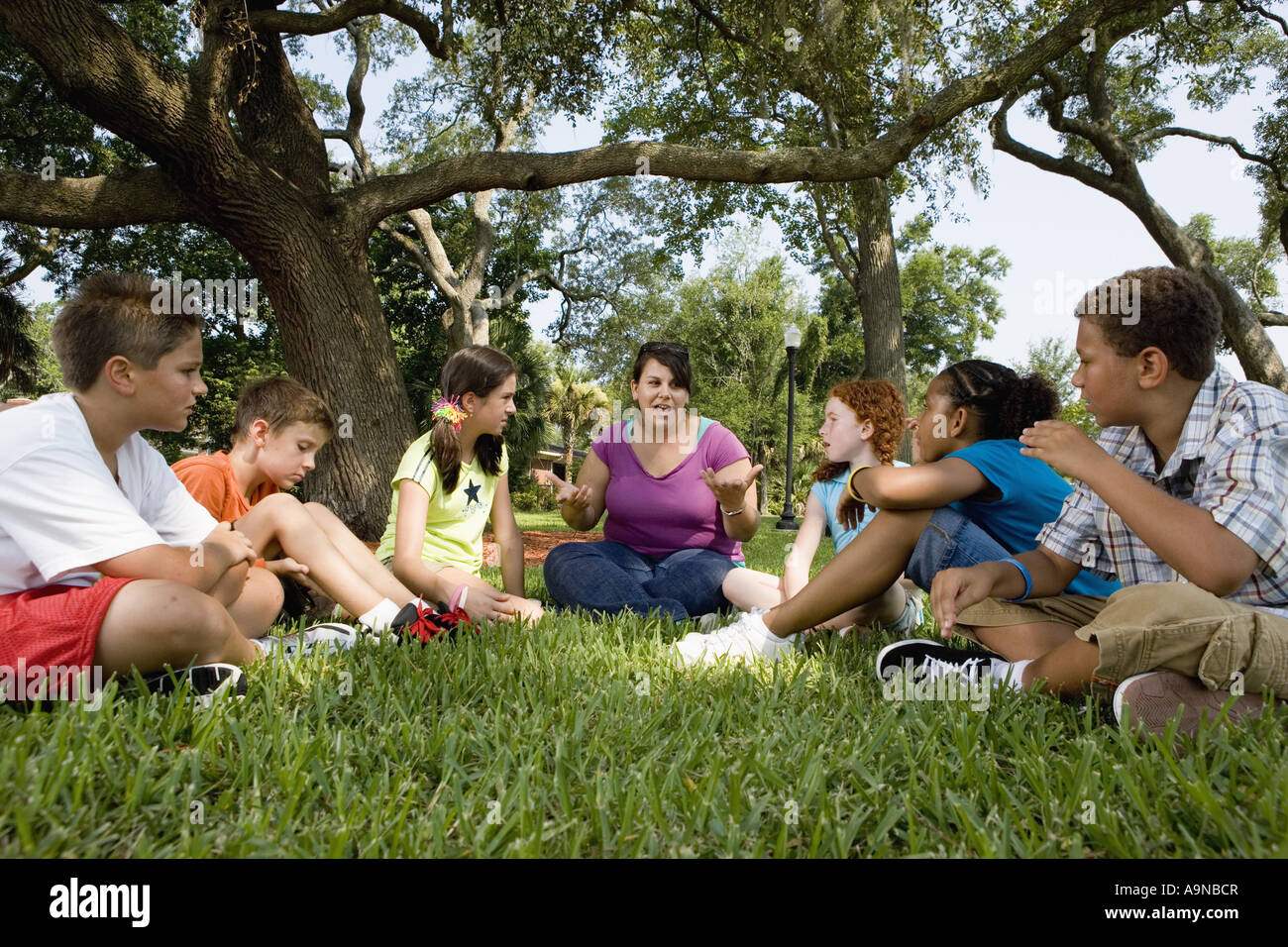 Children sitting in a circle on the grass with their teacher at a park ...