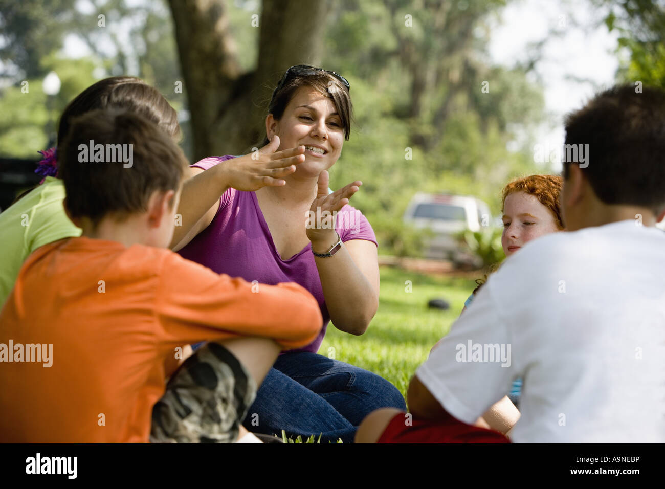 Teacher talking and gesturing to children while seated on the grass at ...
