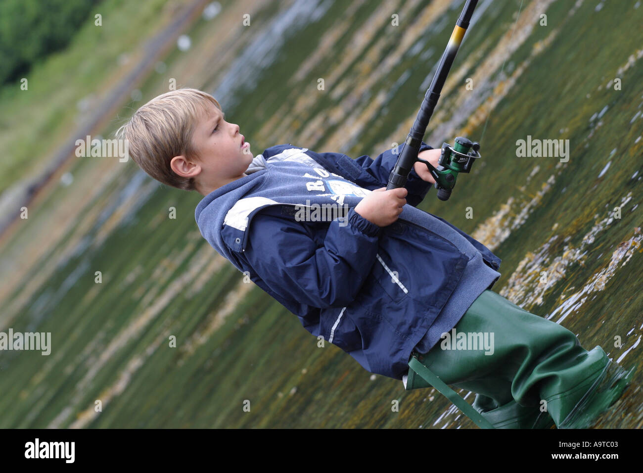Young boy fishing with rod and reel in a Scottish loch wearing wadders ...