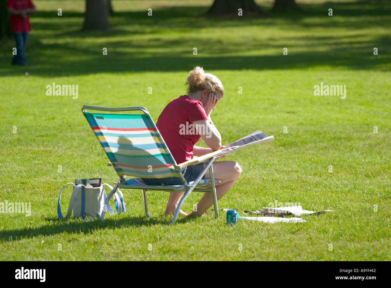 Blond woman sitting on a folding chair reading the newspaper at a park ...