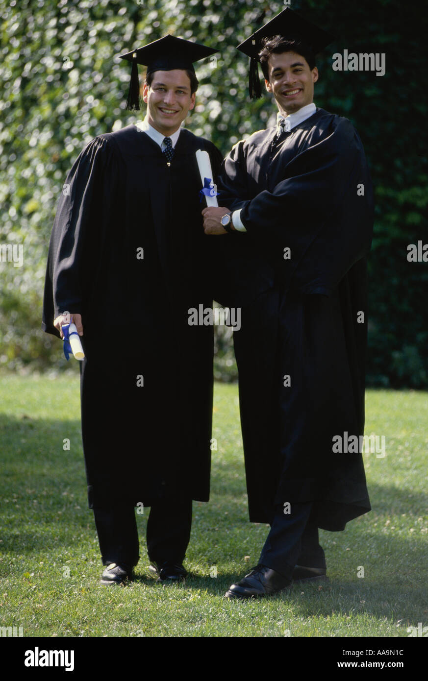 Portrait of two young men wearing graduation outfits holding diplomas ...