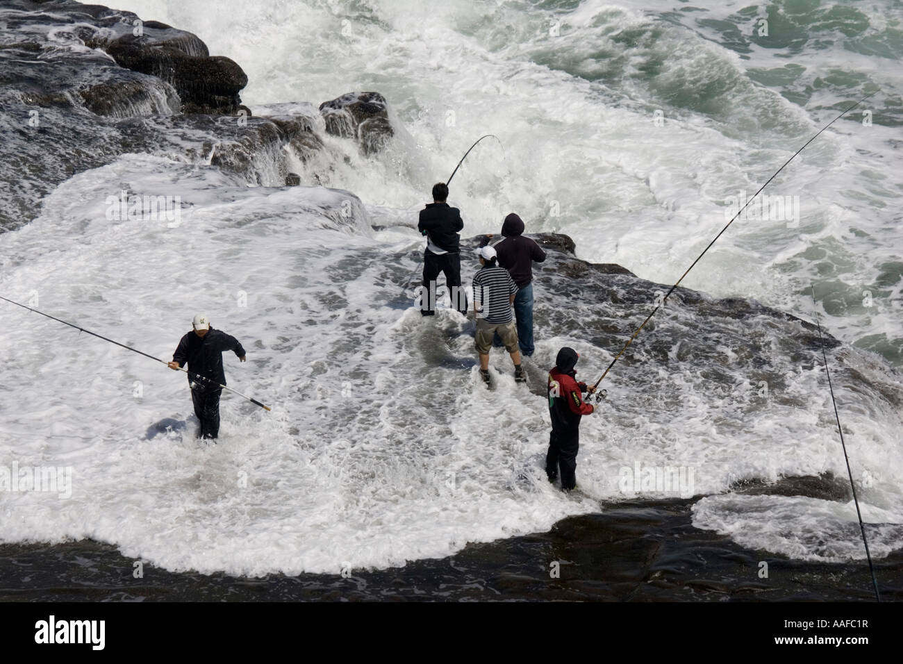 Men fishing on rock outcrop amidst breaking waves on Muriwai beach off ...