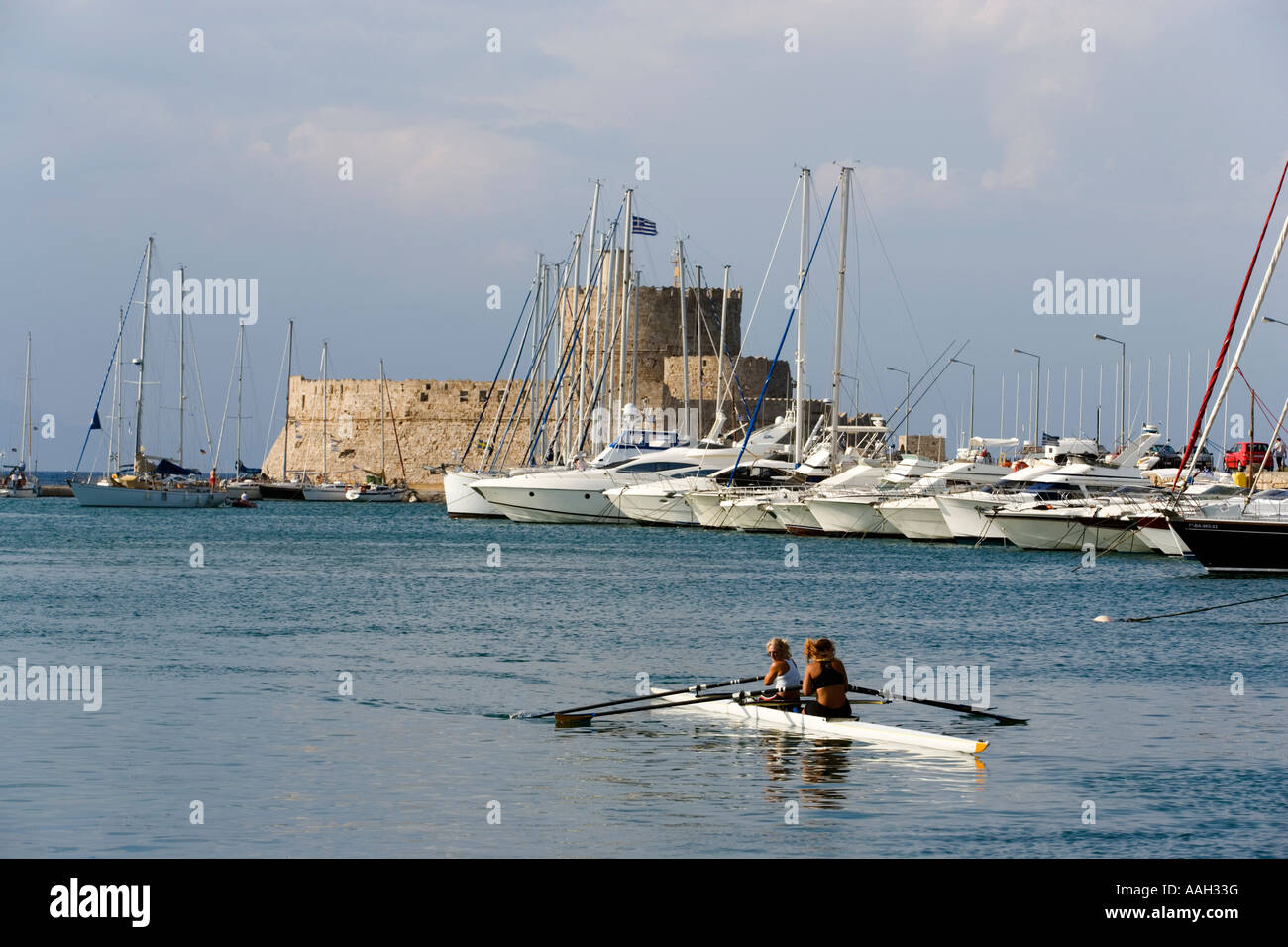 Women rowing in the Mandraki harbour and fortress tower Agios Nikolaos ...