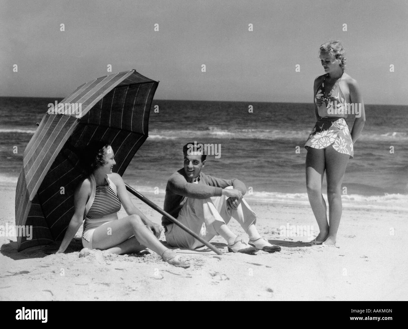 1930s TWO WOMEN 1 MAN SITTING UNDER BEACH UMBRELLA WEARING FASHIONABLE SWIMWEAR Stock Photo - Alamy