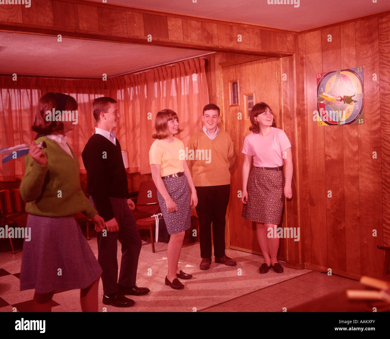 1960s GROUP OF TEENS THROWING DARTS AT DART BOARD IN PANELED REC ROOM ...