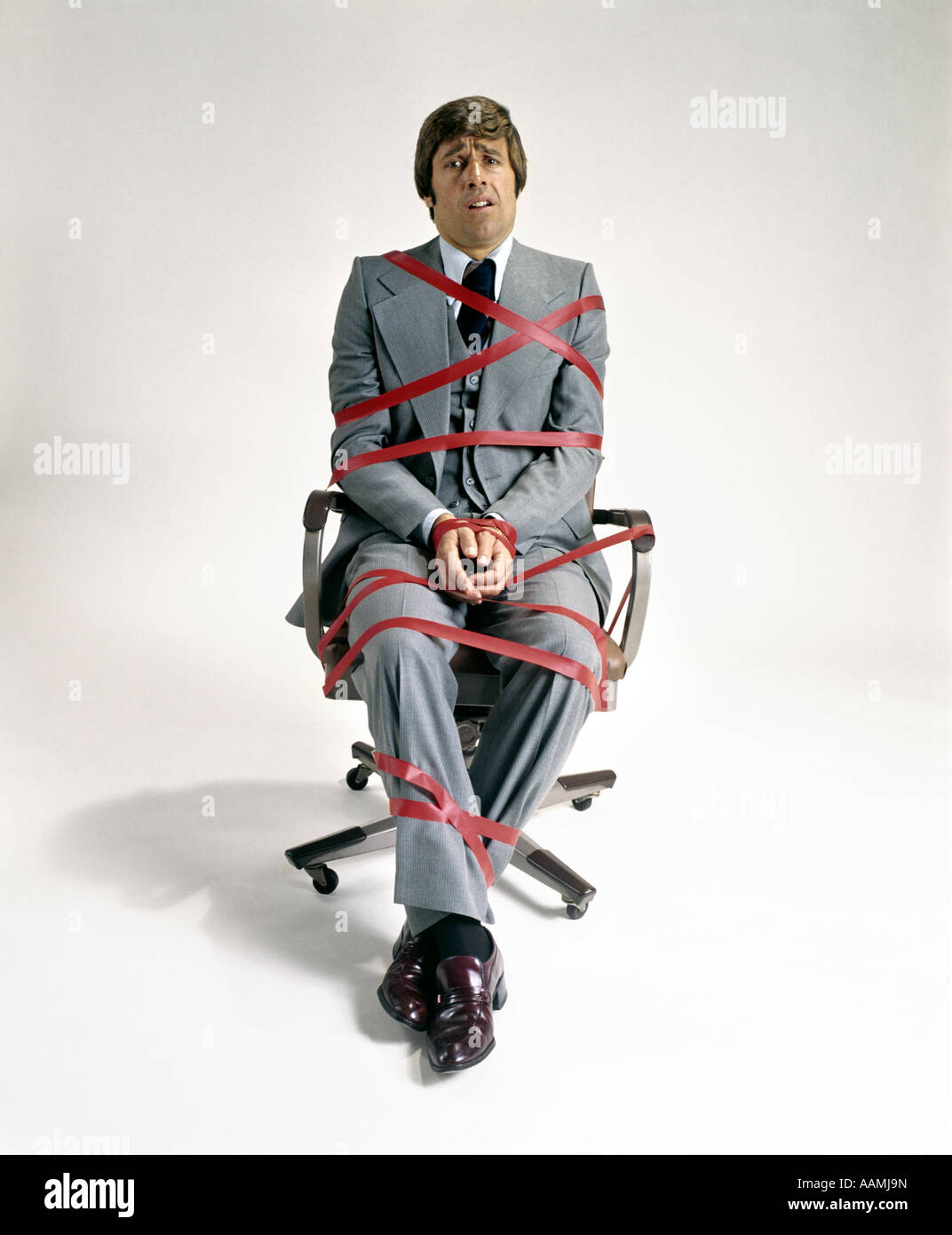 1970s BUSINESS MAN TIED UP IN RED TAPE SITTING IN OFFICE DESK CHAIR ...