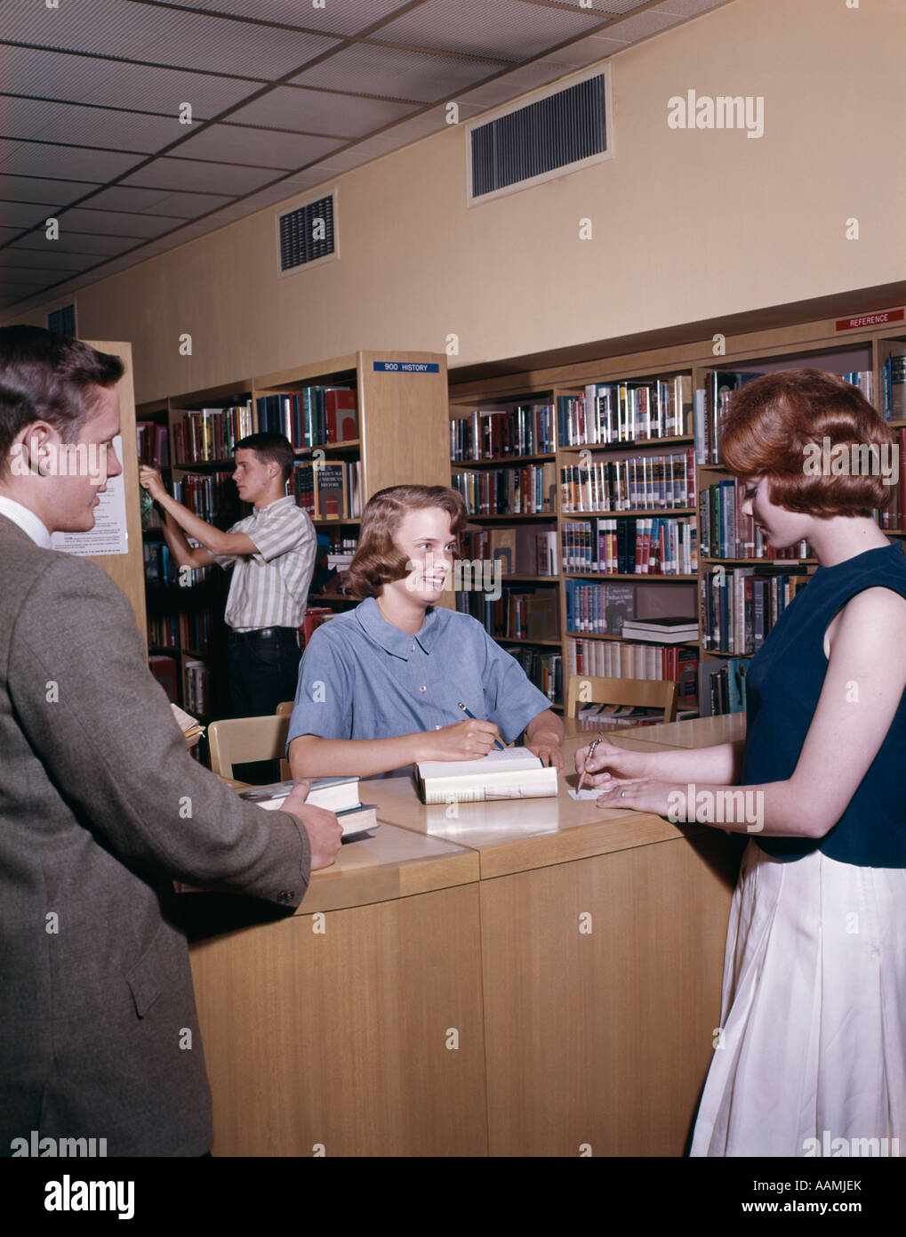1960s FOUR STUDENTS IN COLLEGE LIBRARY THREE AT TABLE AND ONE IN STACKS ...