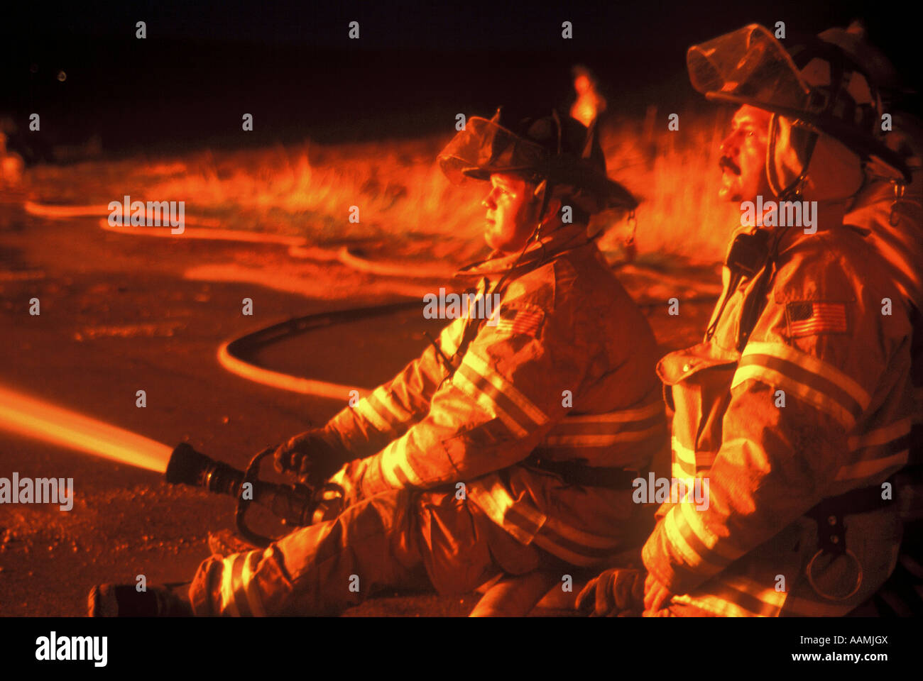 Two fire fighters sitting on a hose putting water on a house on fire ...