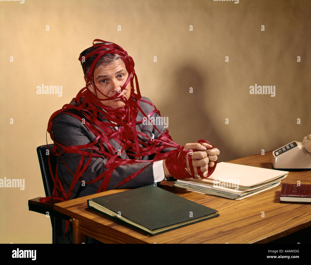 1960s MAN WRAPPED IN RED TAPE SITTING AT DESK WITH HANDS ON TOP OF ...