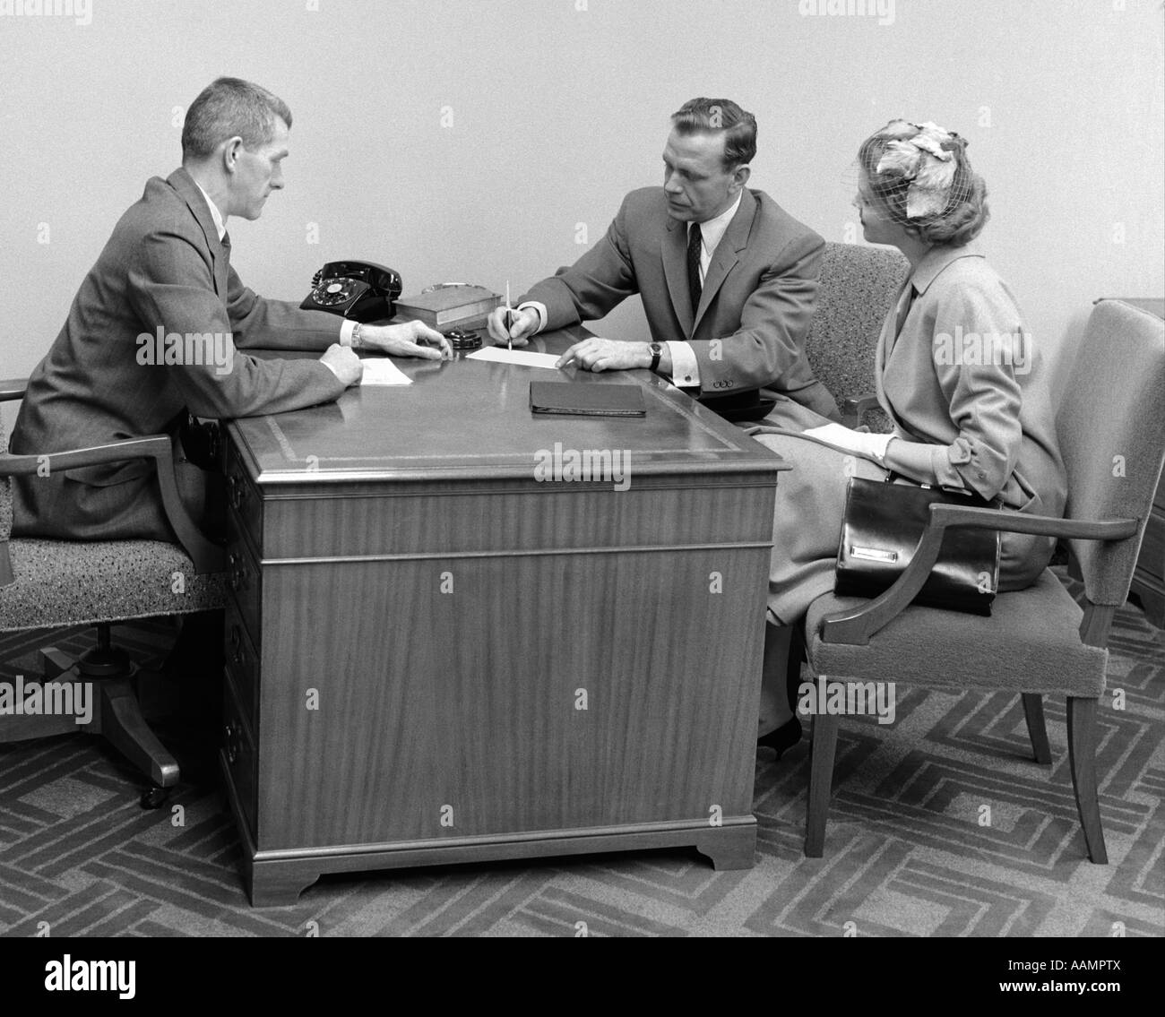 1960s SIDE VIEW OF COUPLE SITTING AT DESK WITH EXECUTIVE SIGNING PAPERS ...