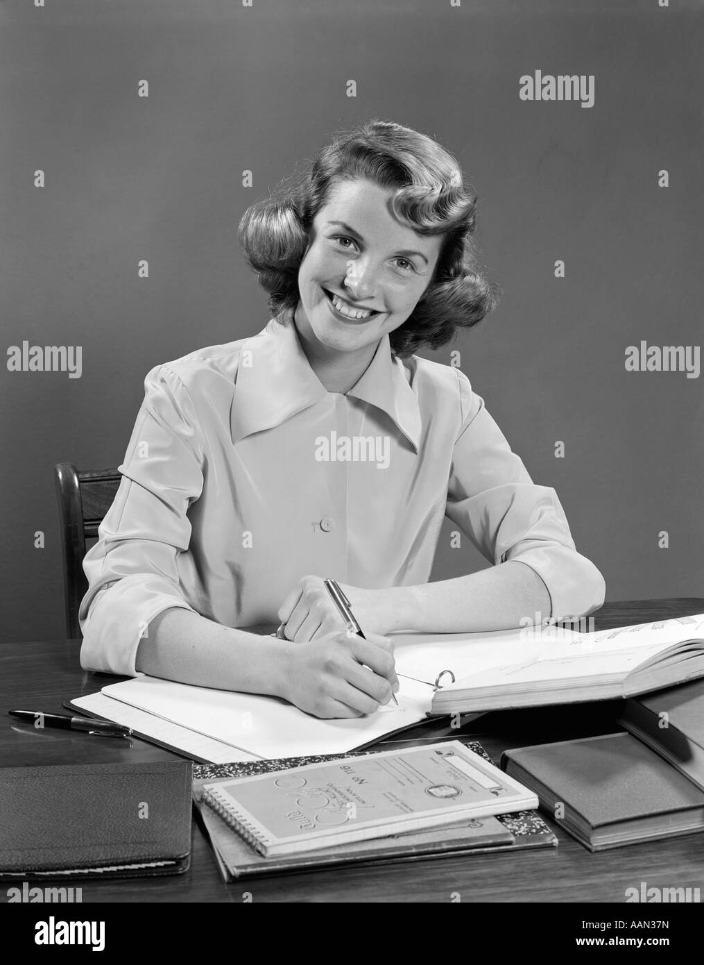 1950s SMILING YOUNG WOMAN SITTING AT DESK WRITING AND STUDYING LOOKING ...