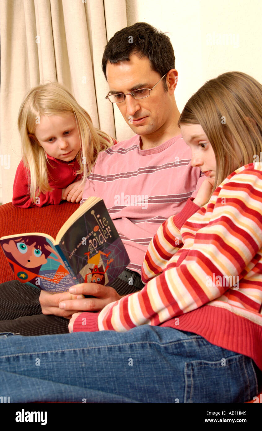 Father reading book to his daughters on sofa in living room at home UK ...