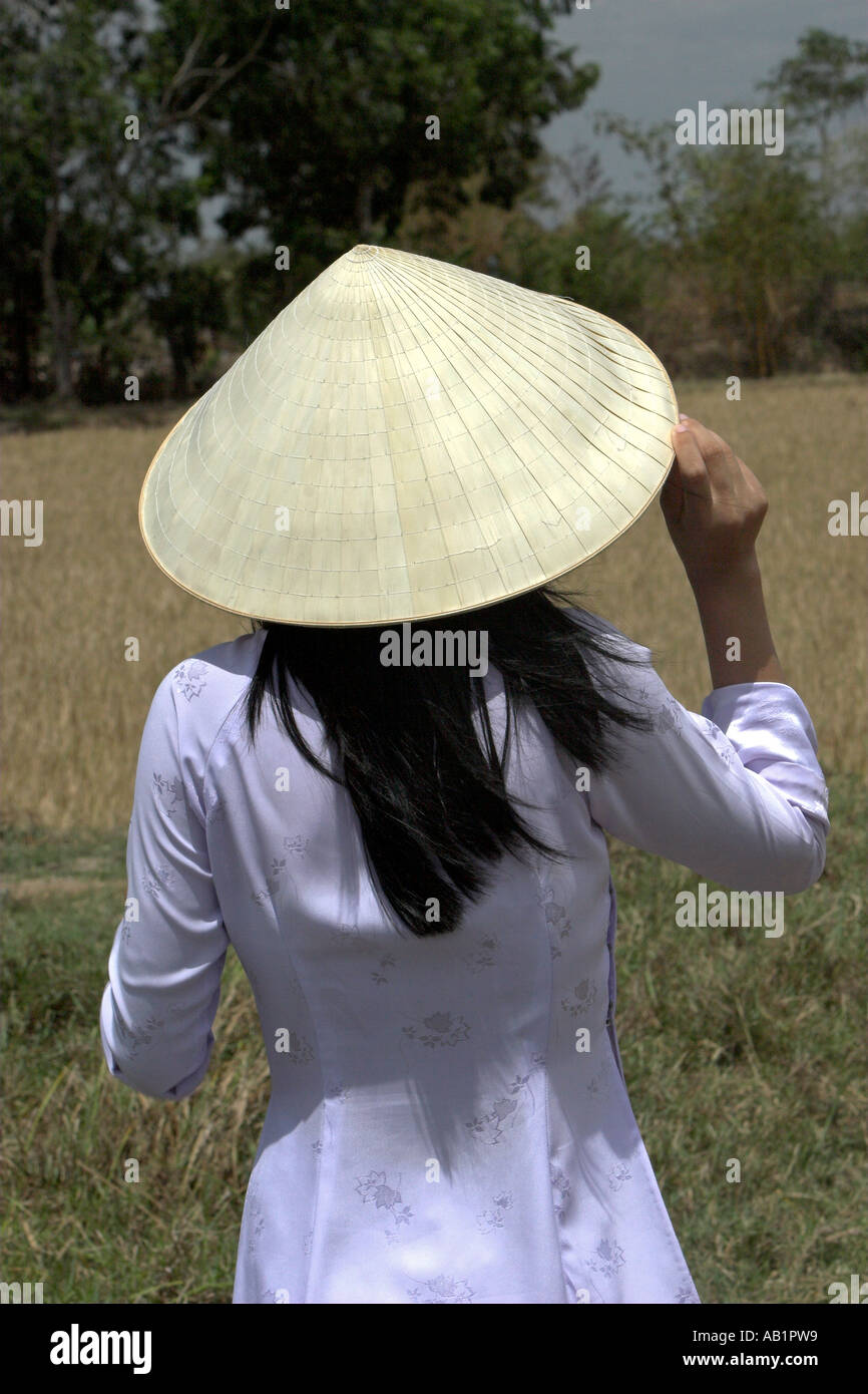 Young woman in traditional white ao dai holding conical hat on head looks across fields Vietnam ...
