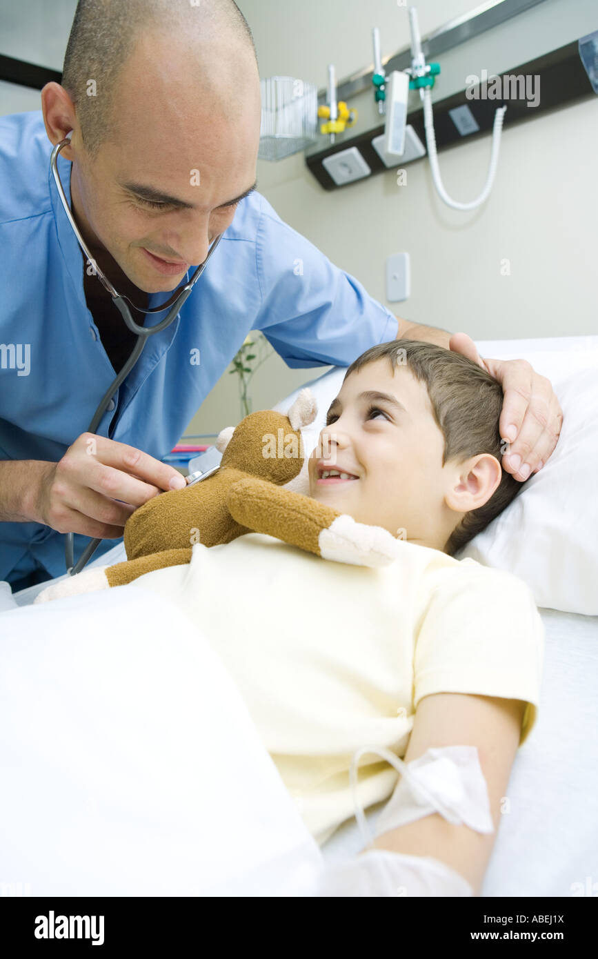 Boy lying in hospital bed, intern holding stethoscope to boy's stuffed ...