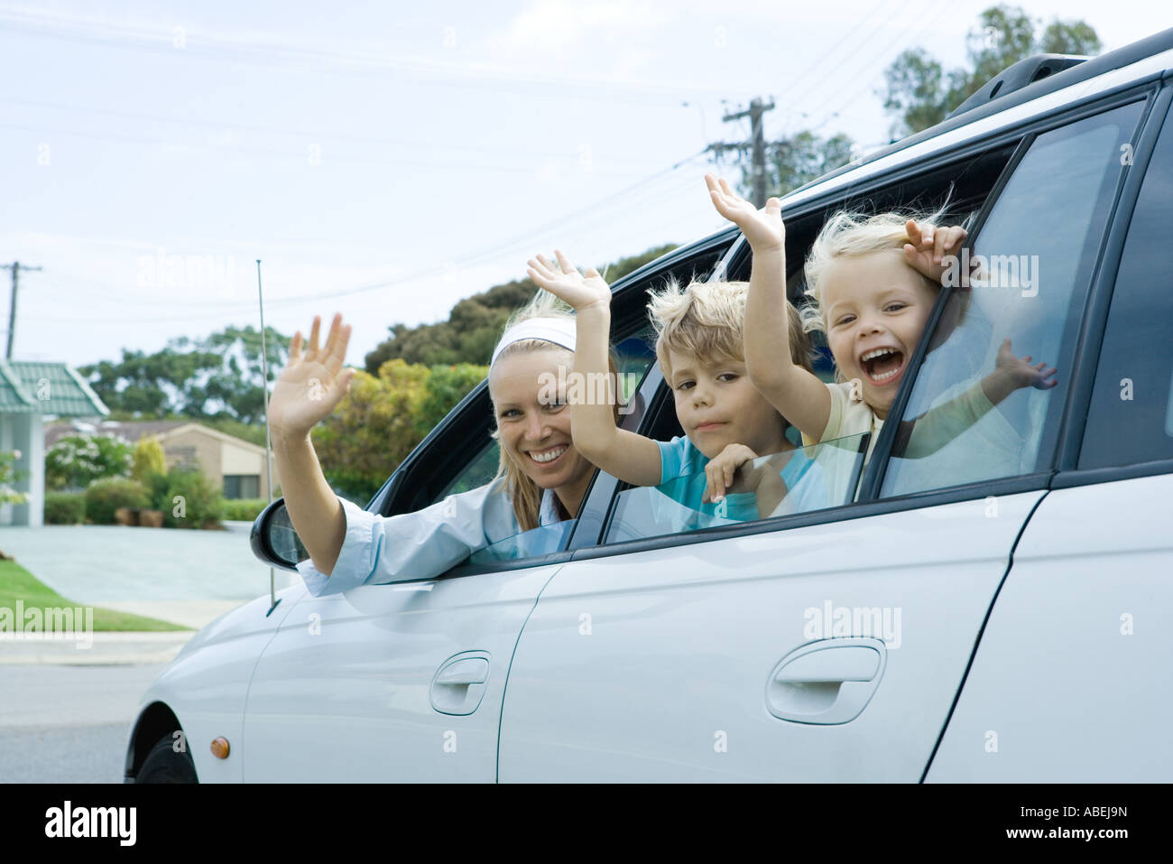 Mother and two children waving out of car windows, looking at camera ...