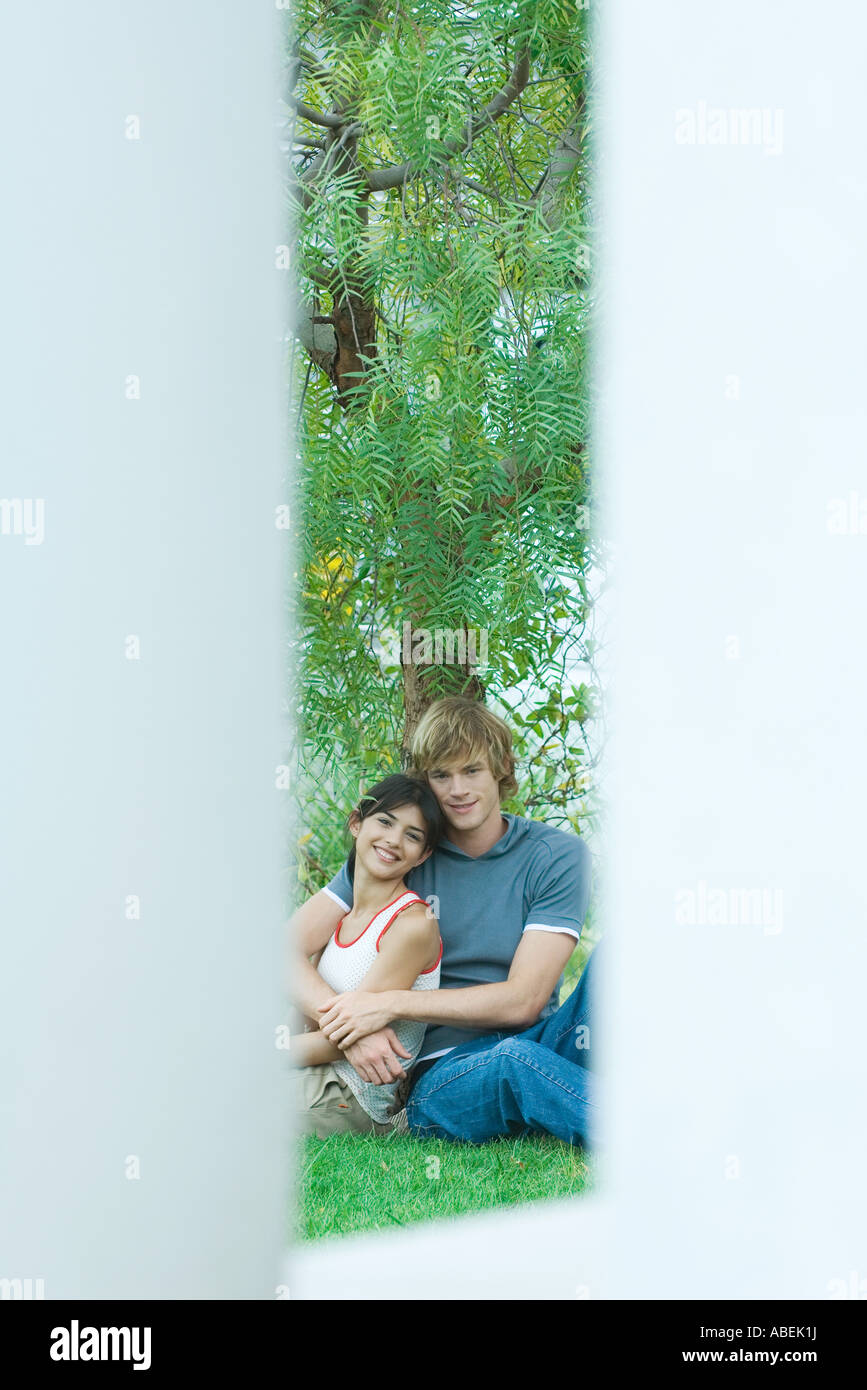Young couple sitting on ground, leaning against tree, smiling at camera ...