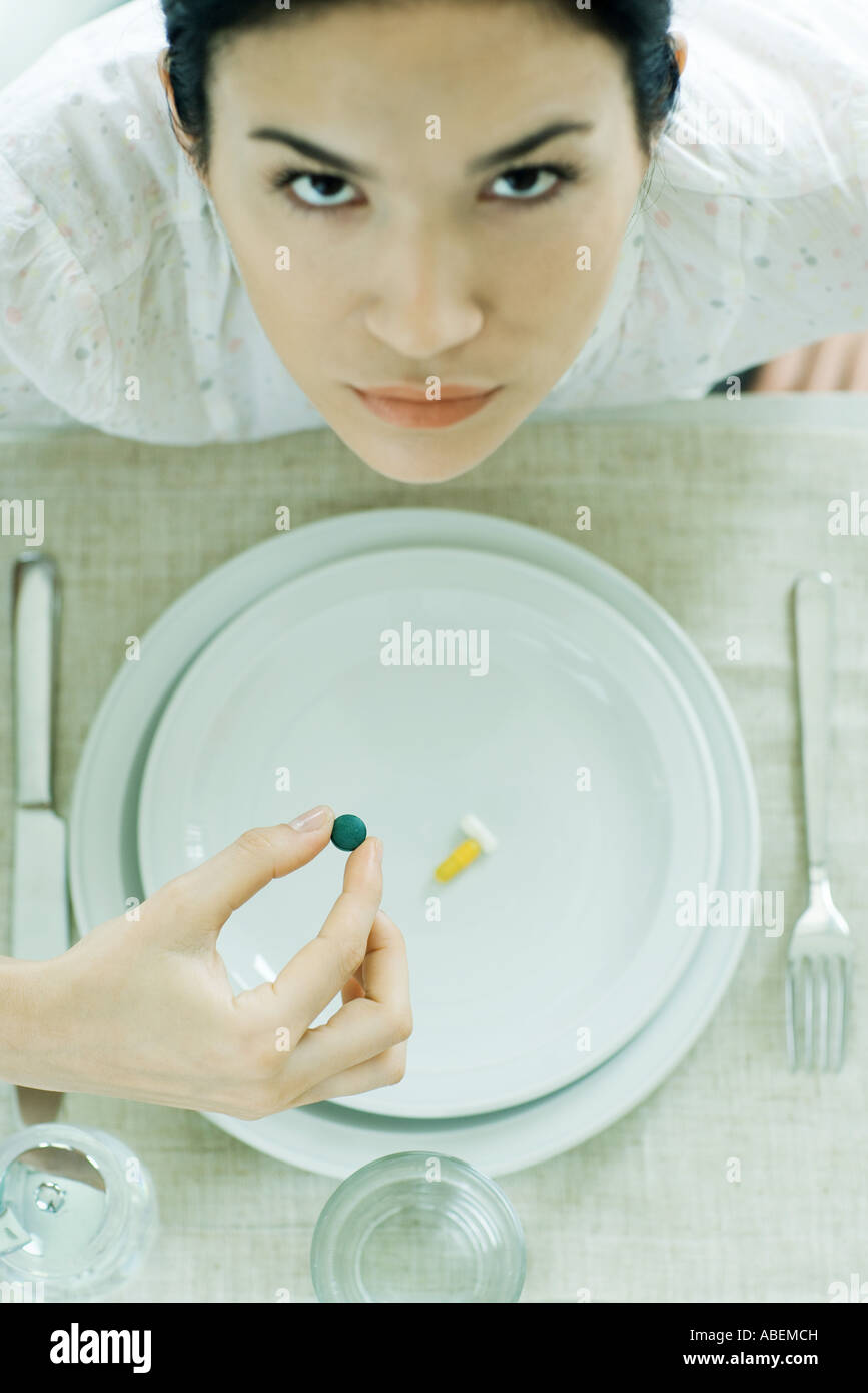Woman sitting at empty plate holding vitamin, looking up at camera ...