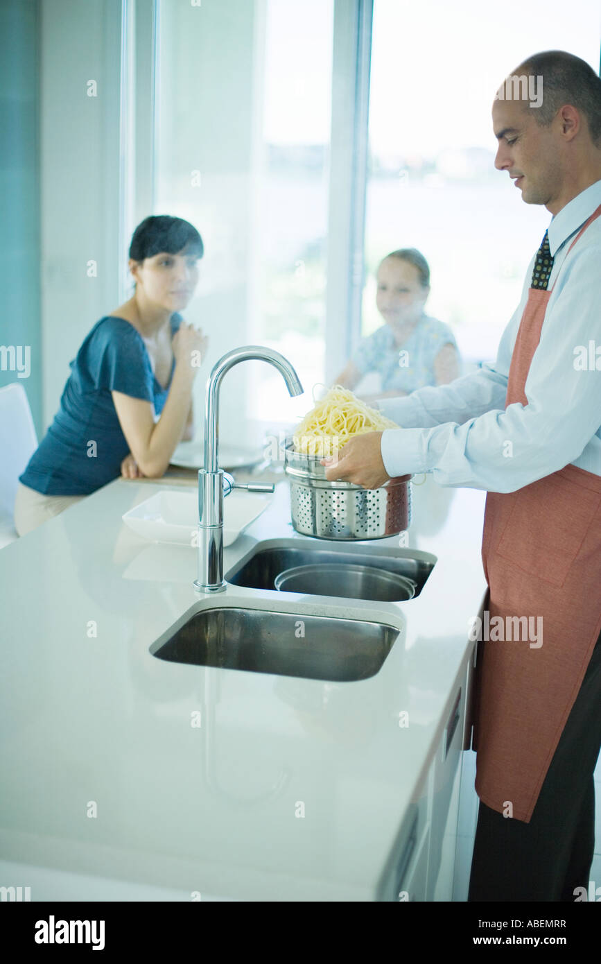 Man draining spaghetti over kitchen sink while wife and daughter watch ...