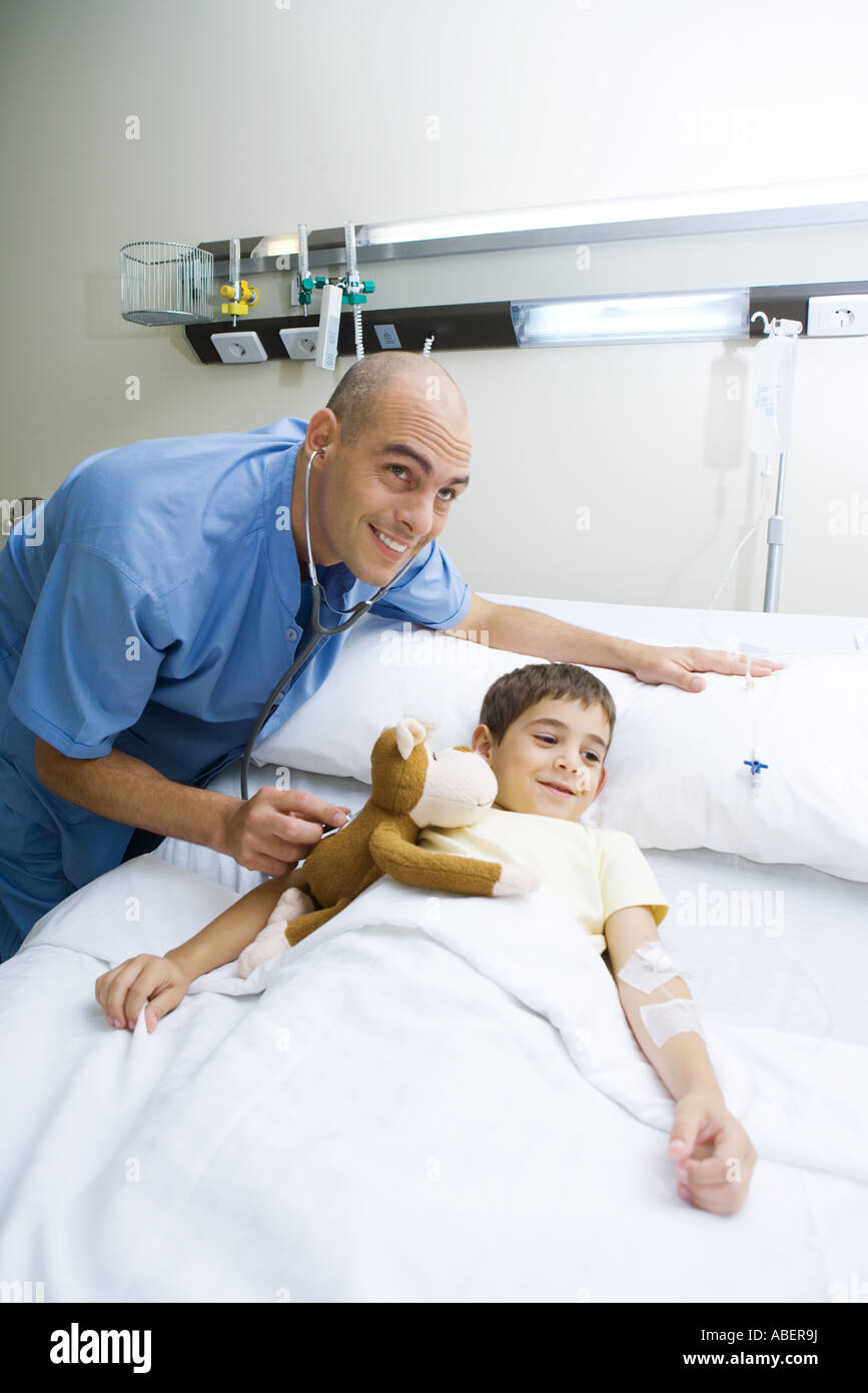 Boy lying in hospital bed, doctor holding stethoscope to stuffed monkey ...
