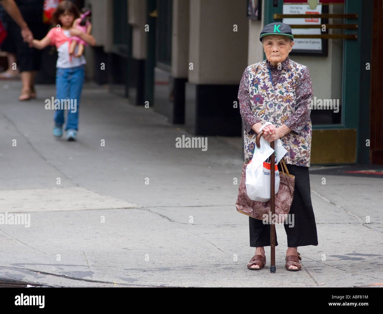 An elderly Asian woman waits to cross the street at a crosswalk while a ...