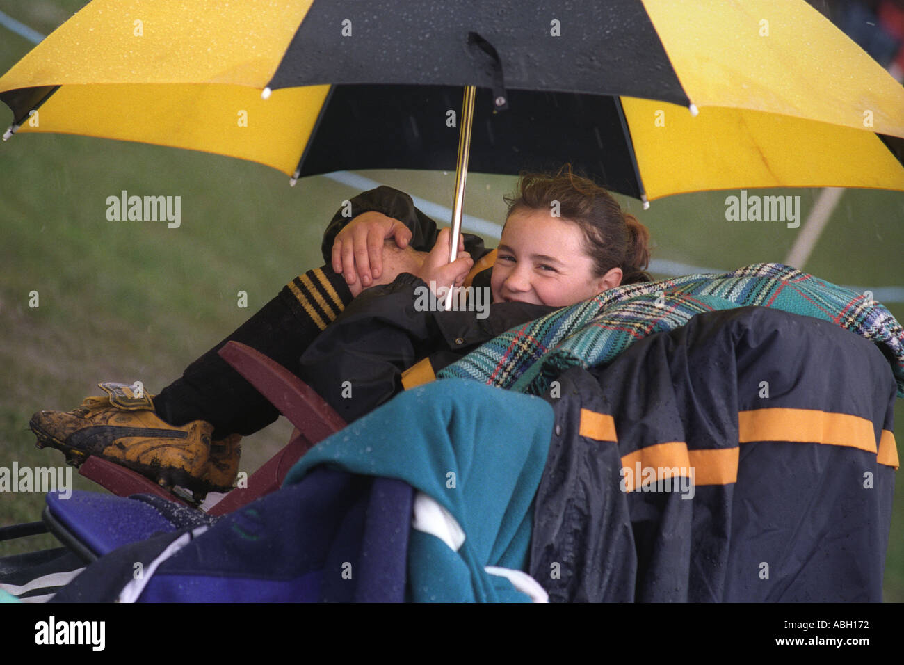 Young girl wearing rugby boots sheltering under an umbrella during a ...