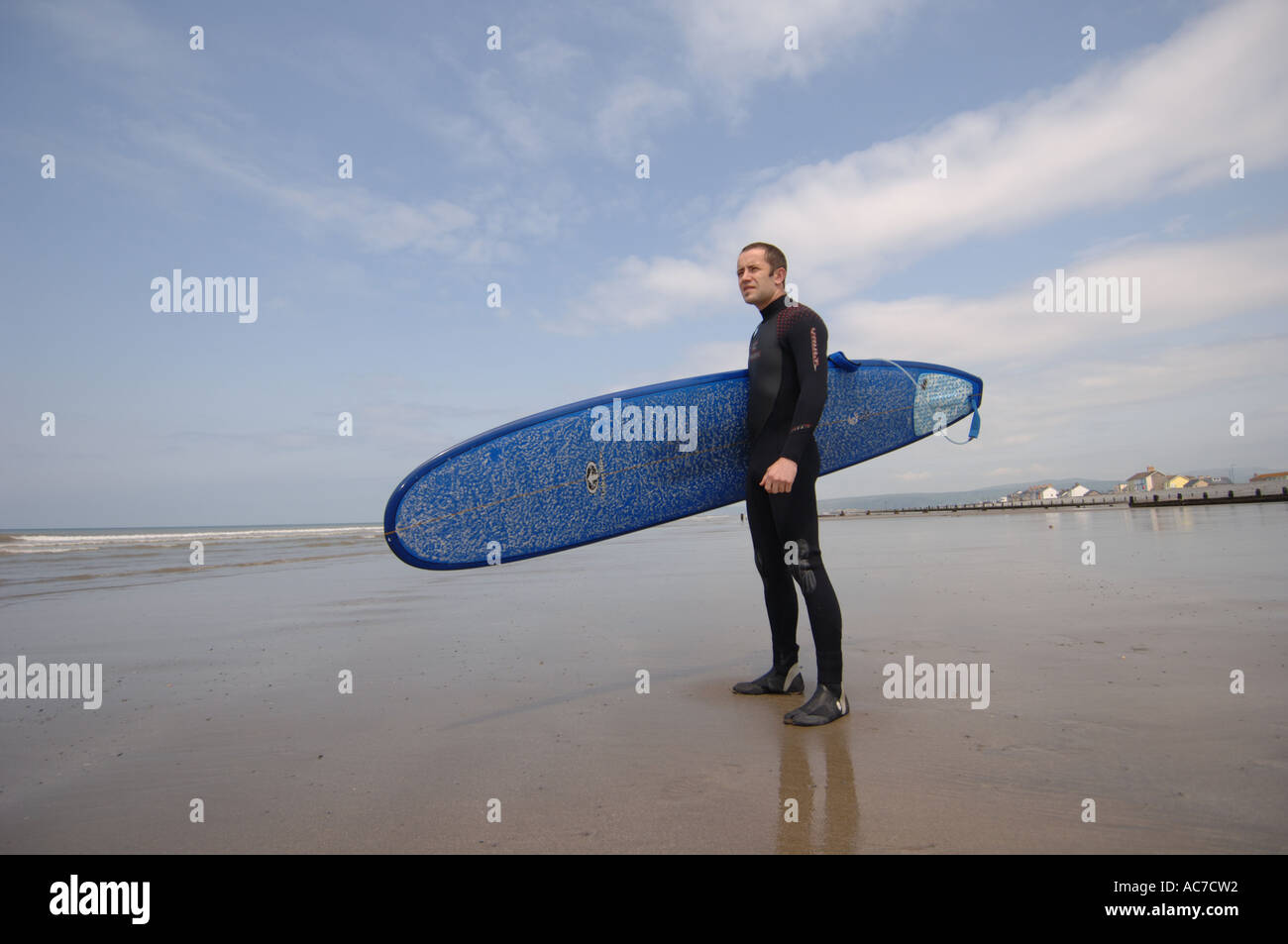 A male surfer wearing a wetsuit holding his surf board watches for ...