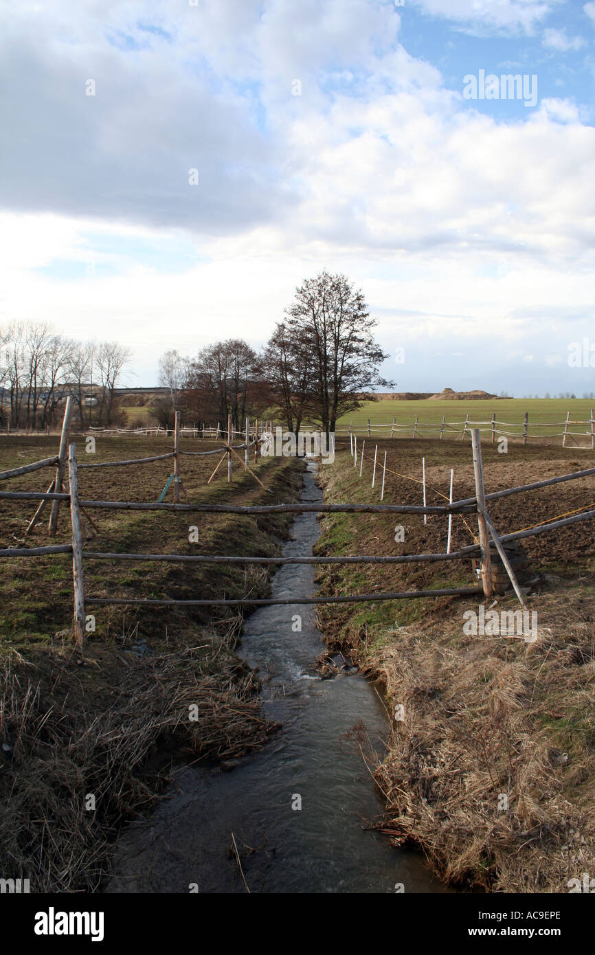Small stream flowing through a rural field with wooden fences and trees ...