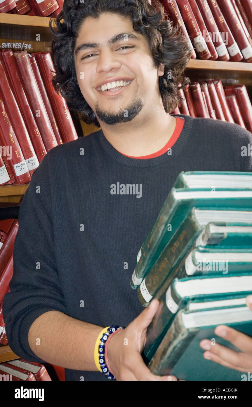 Portrait of a young man carrying a stack of books in a library and ...