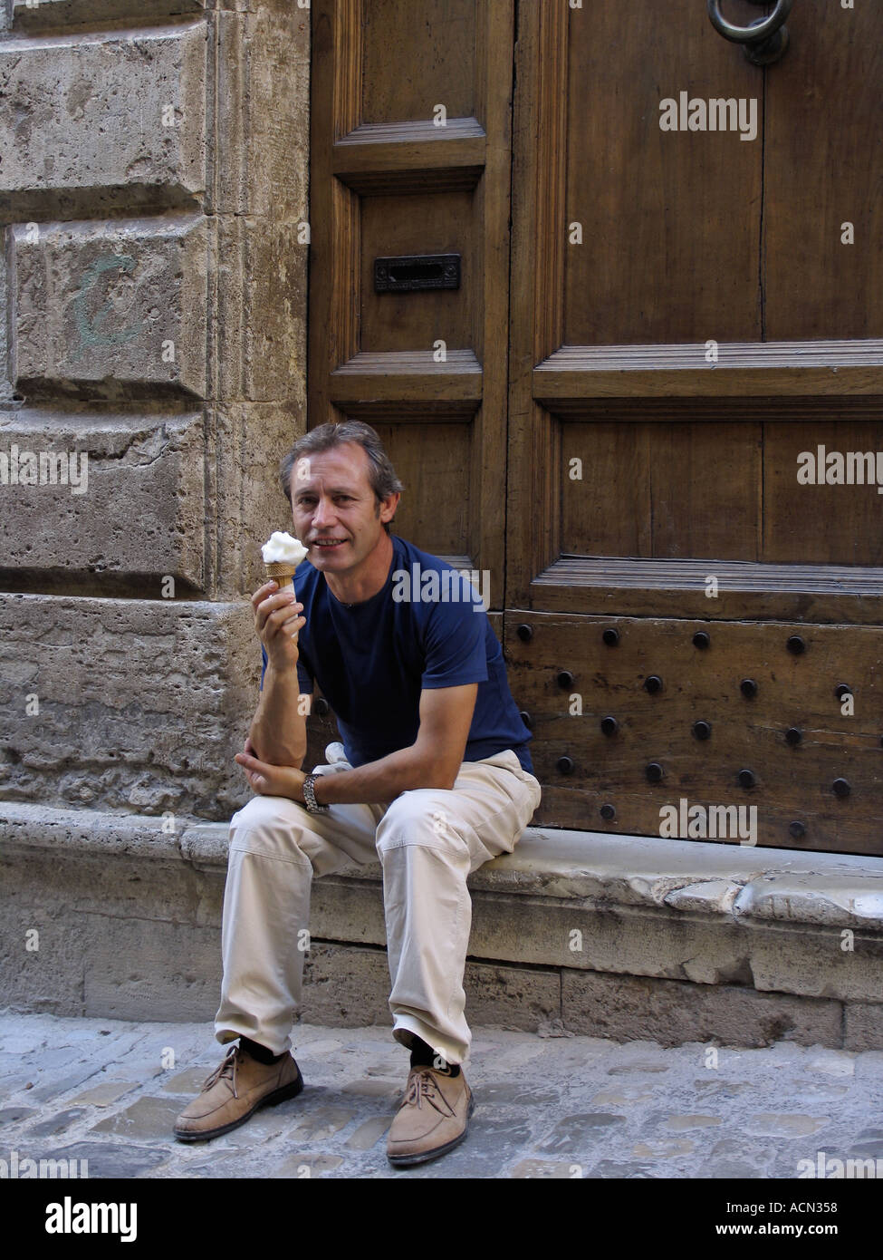 Italian man eating gelato cone sits on ledge beside stone wall in front ...