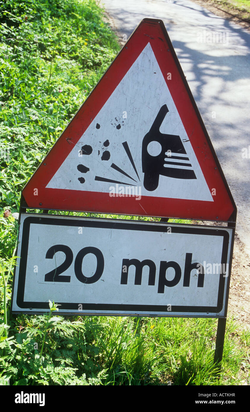 Red white and black sign on grass verge of narrow lane warning of loose ...