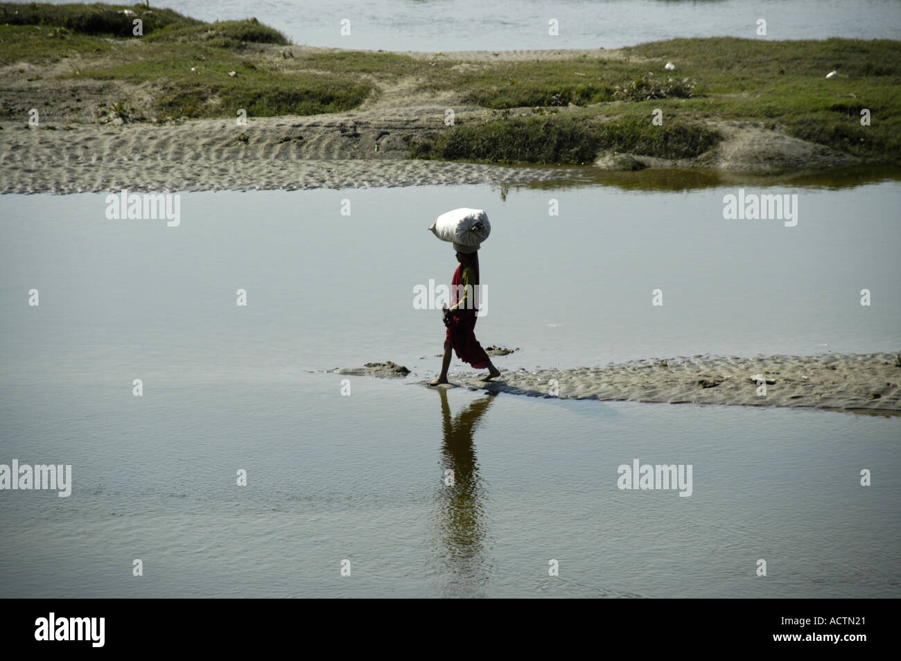 Woman carrying a sack on her head is walking into the water of river ...