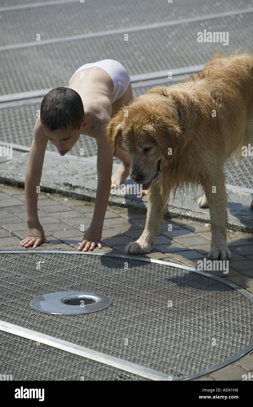 A young boy mimics a dog as it waits for a fountain to spout water ...