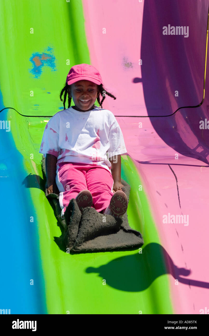 Black girl in pink outfit smiling as she slides down a colourful slide ...