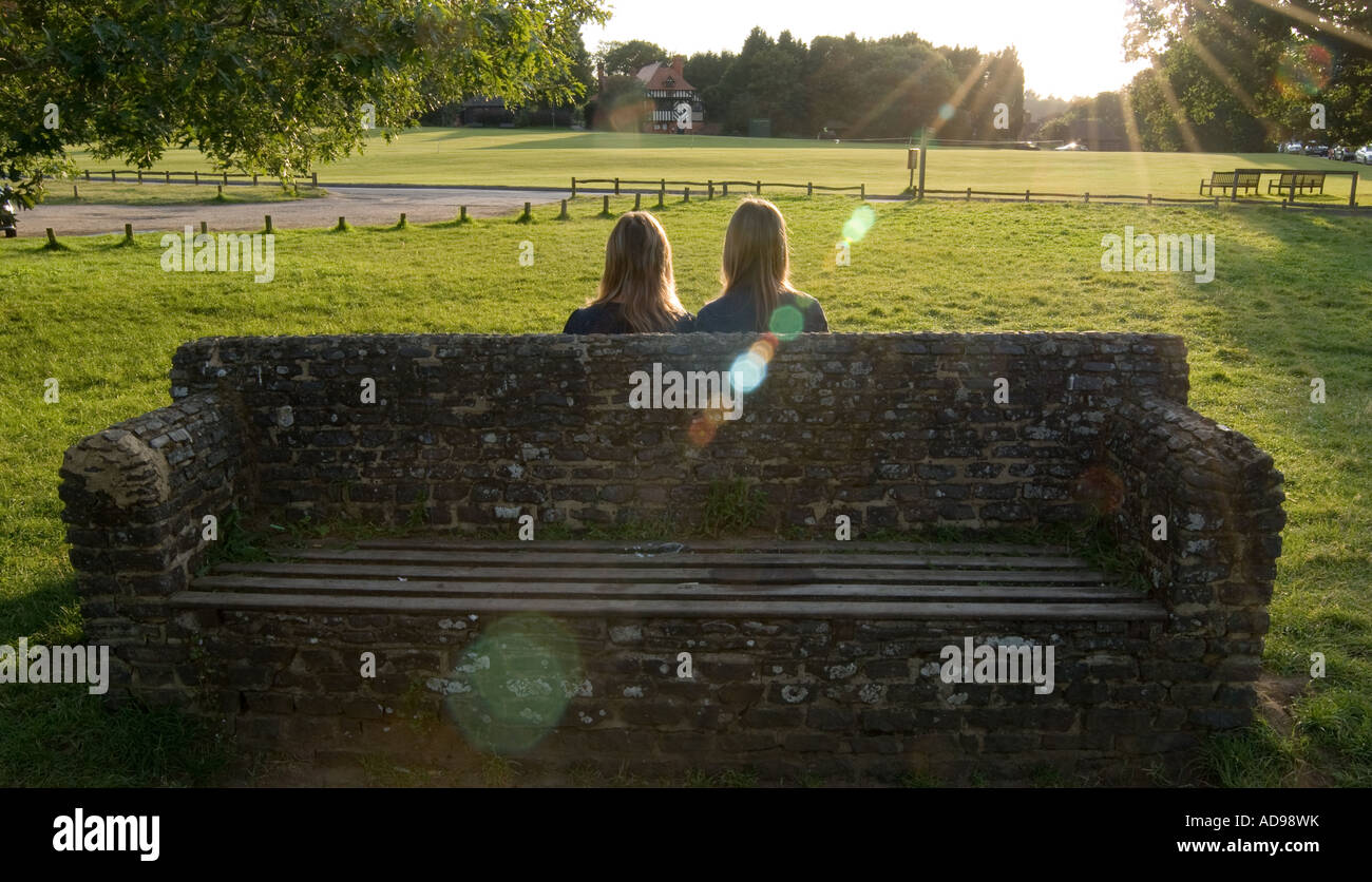 Two women sit alone on a stone bench facing away looking into the light ...