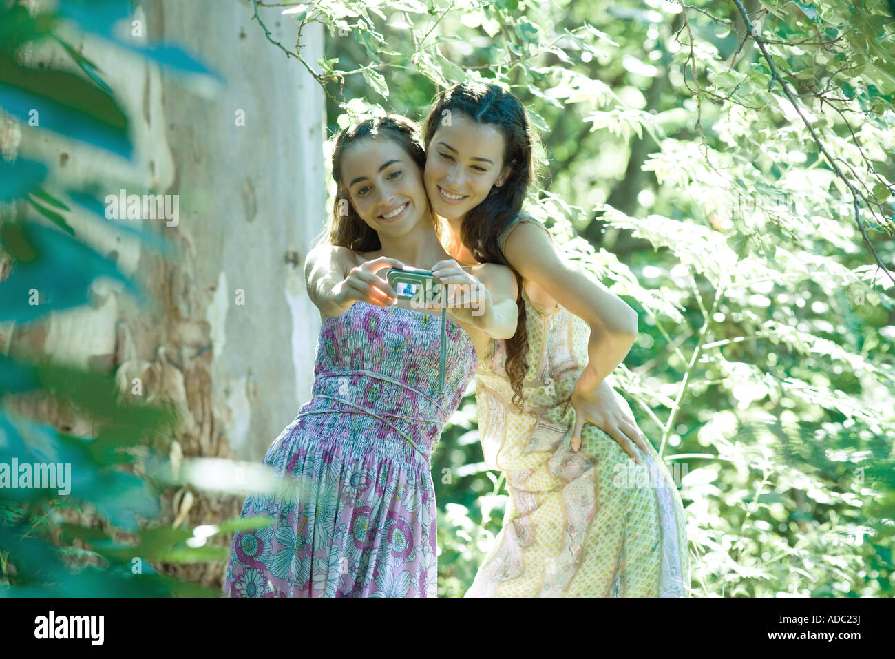 Two young women wearing sundresses, standing in forest, taking photo of ...