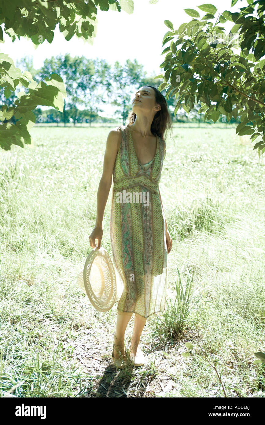 Young woman wearing sundress and holding sun hat, standing looking up ...