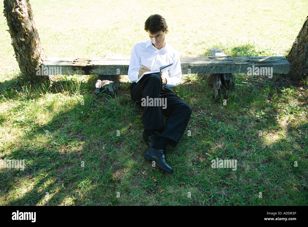 Businessman sitting on ground, leaning against bench, writing Stock ...