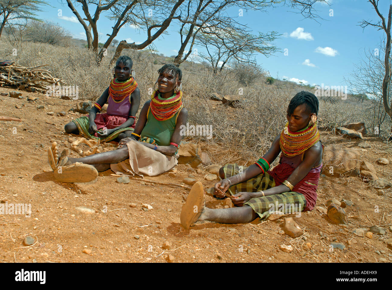 Three Turkana women with colorful bead necklaces sitting on a dirt road ...