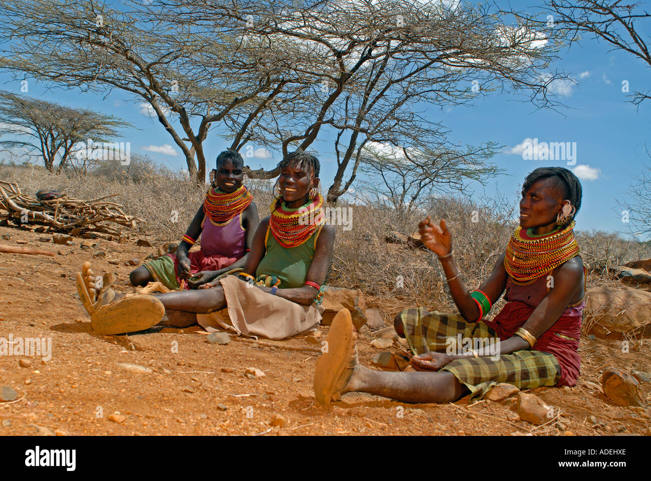 Three Turkana women with colorful bead necklaces sitting on a dirt road ...