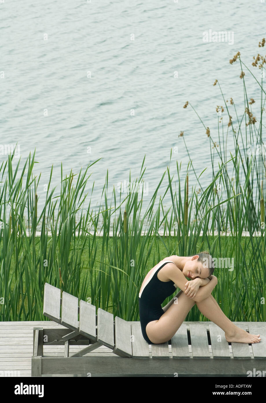 Woman sitting on lounge chair next to lake, hugging knees, resting head ...