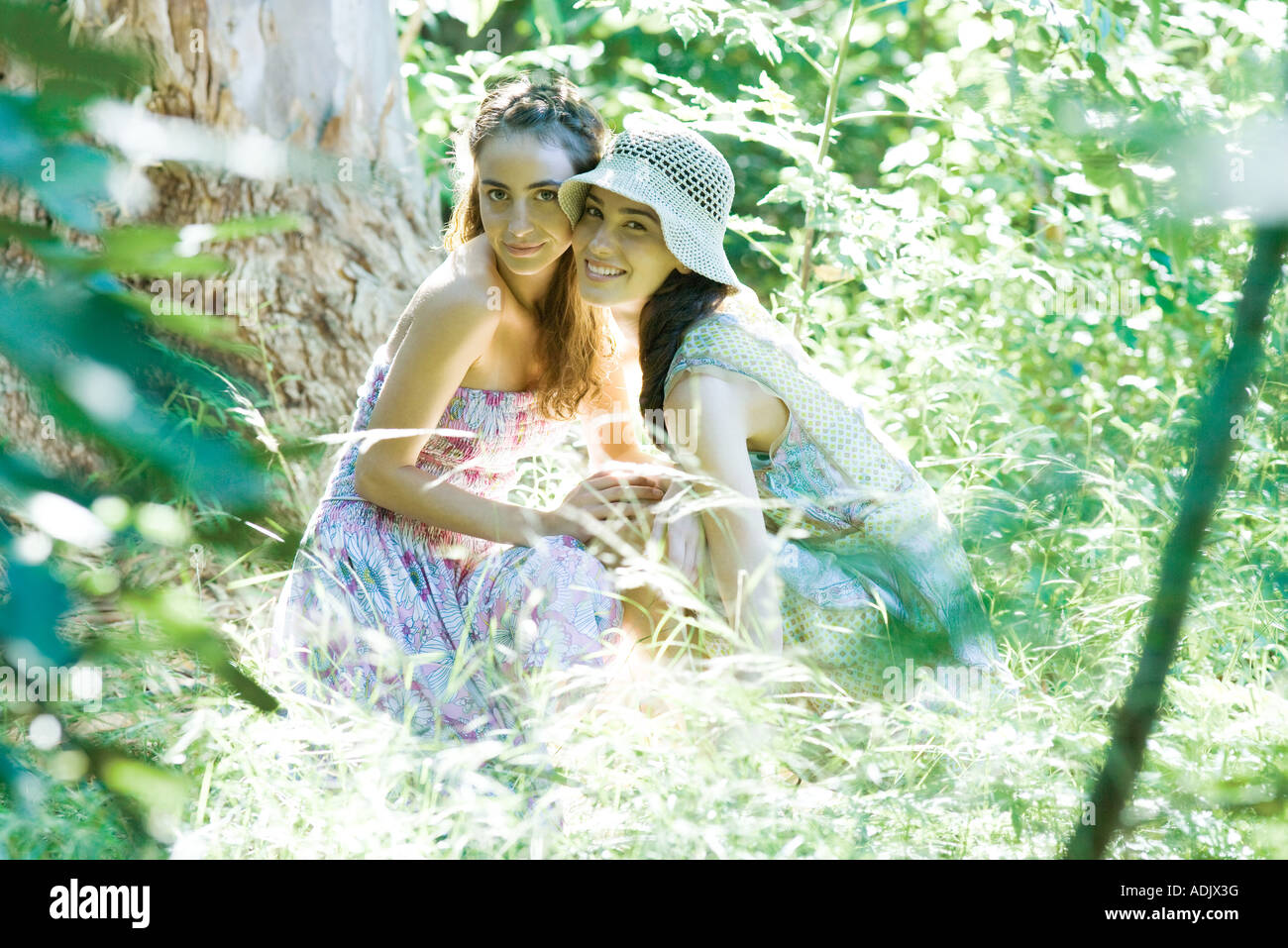 Two young women wearing sun dresses, crouching near tree with cheeks ...