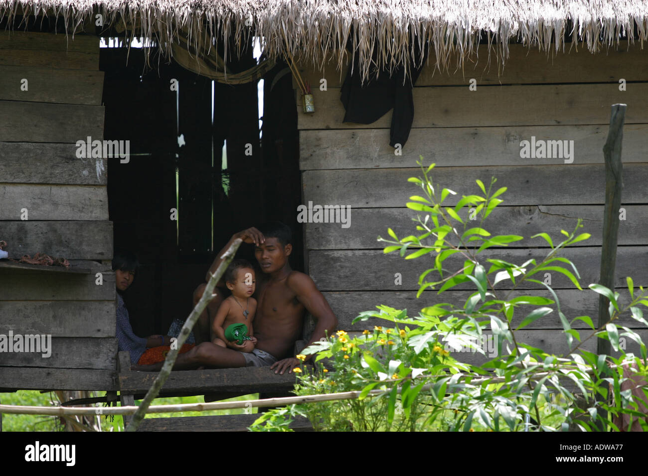 A rural Cambodian farming family sit in the doorway of their wooden hut ...