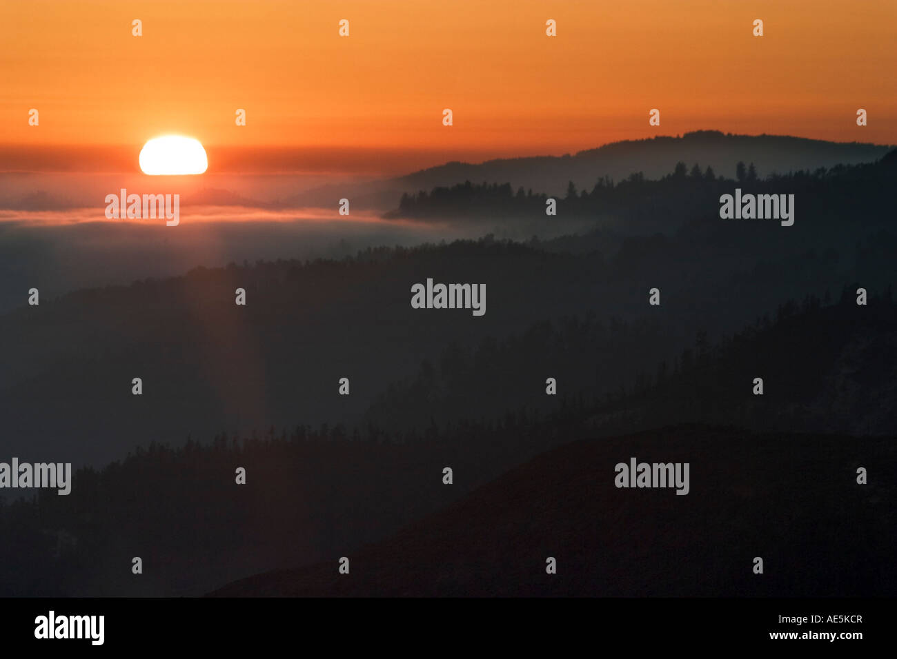 Sun setting over layers of mountains with fog amid glowing orange sky ...