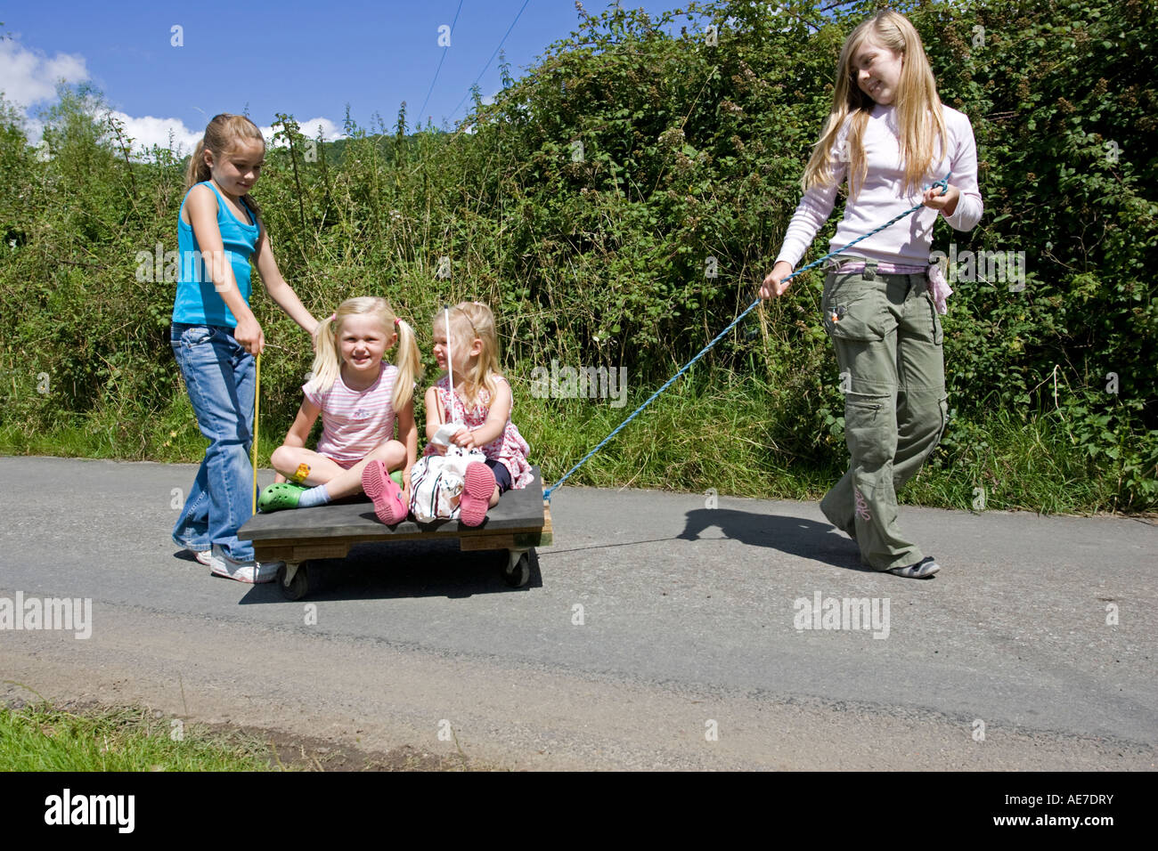 Girls having fun pulling younger sisters along lane on homemade trolley ...