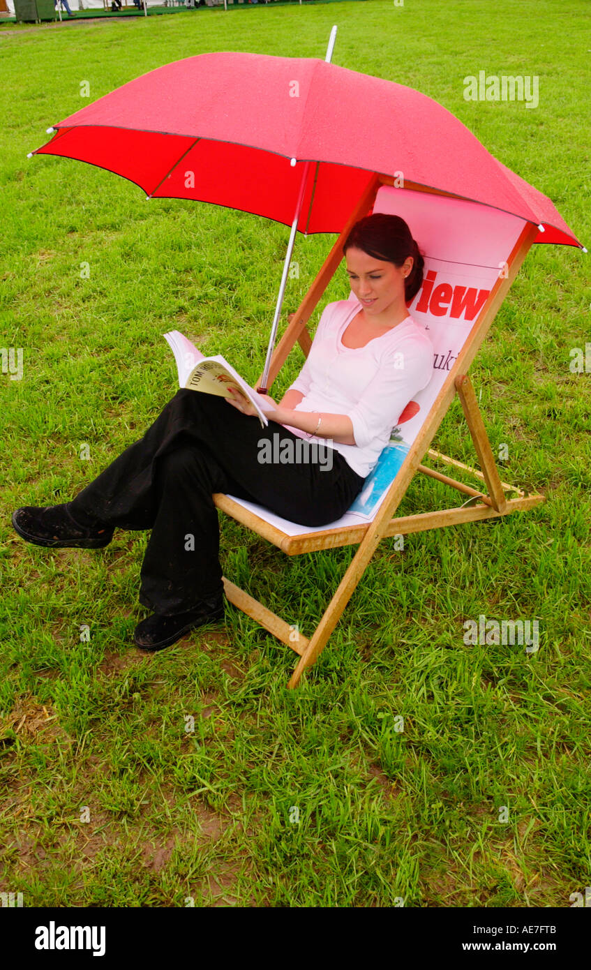 Young woman reading in rain sat in deckchair under red umbrella at The ...