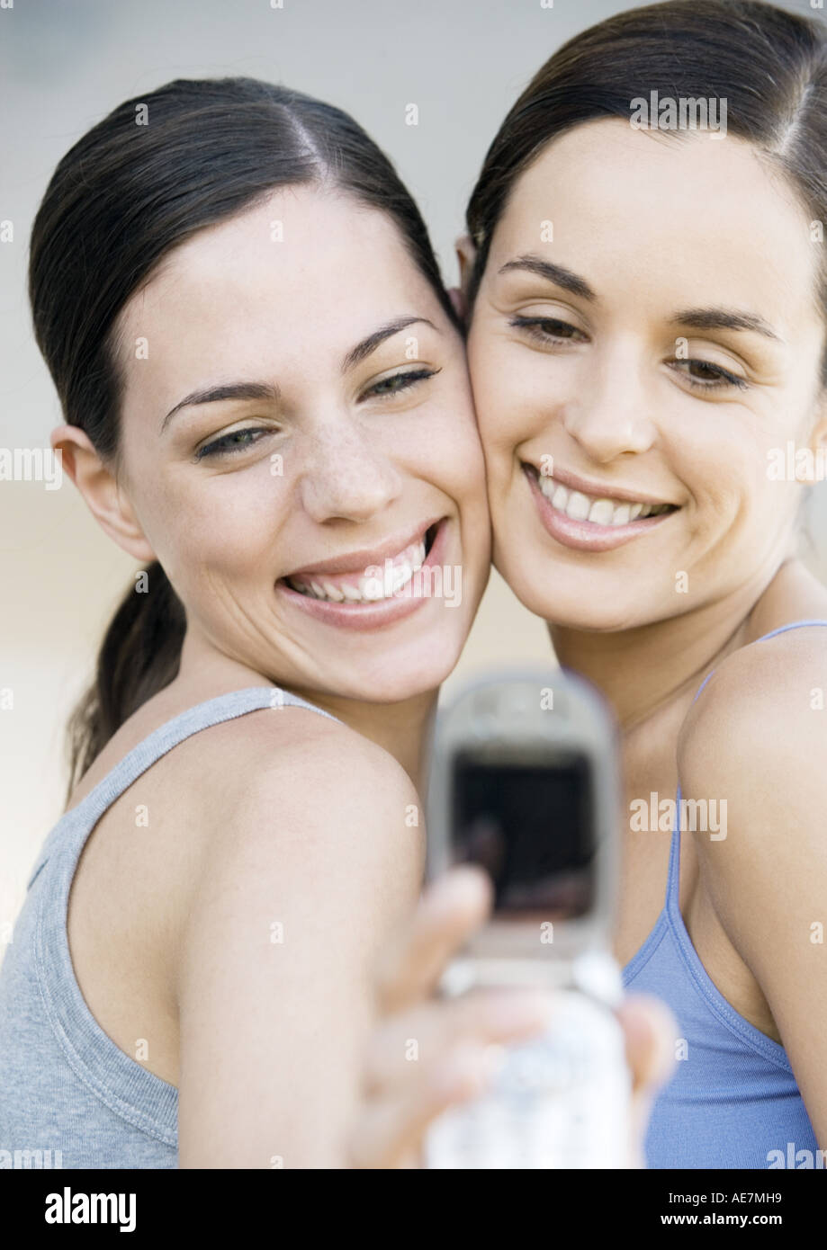 Two young female friends, cheek to cheek, taking photo with cell phone ...