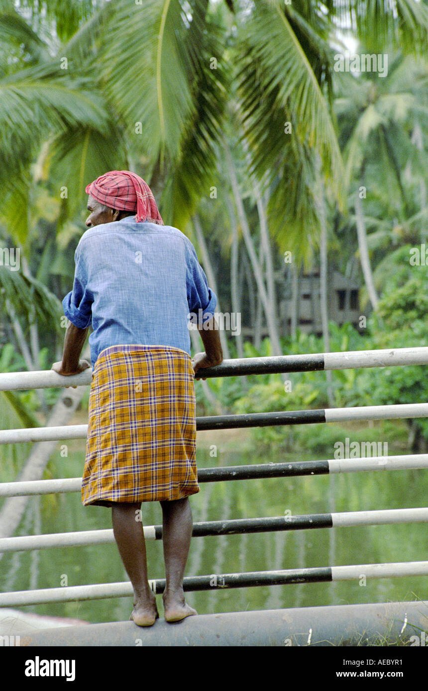 An old manual labourer relaxing on a bridge in Kerala with coconut tree ...